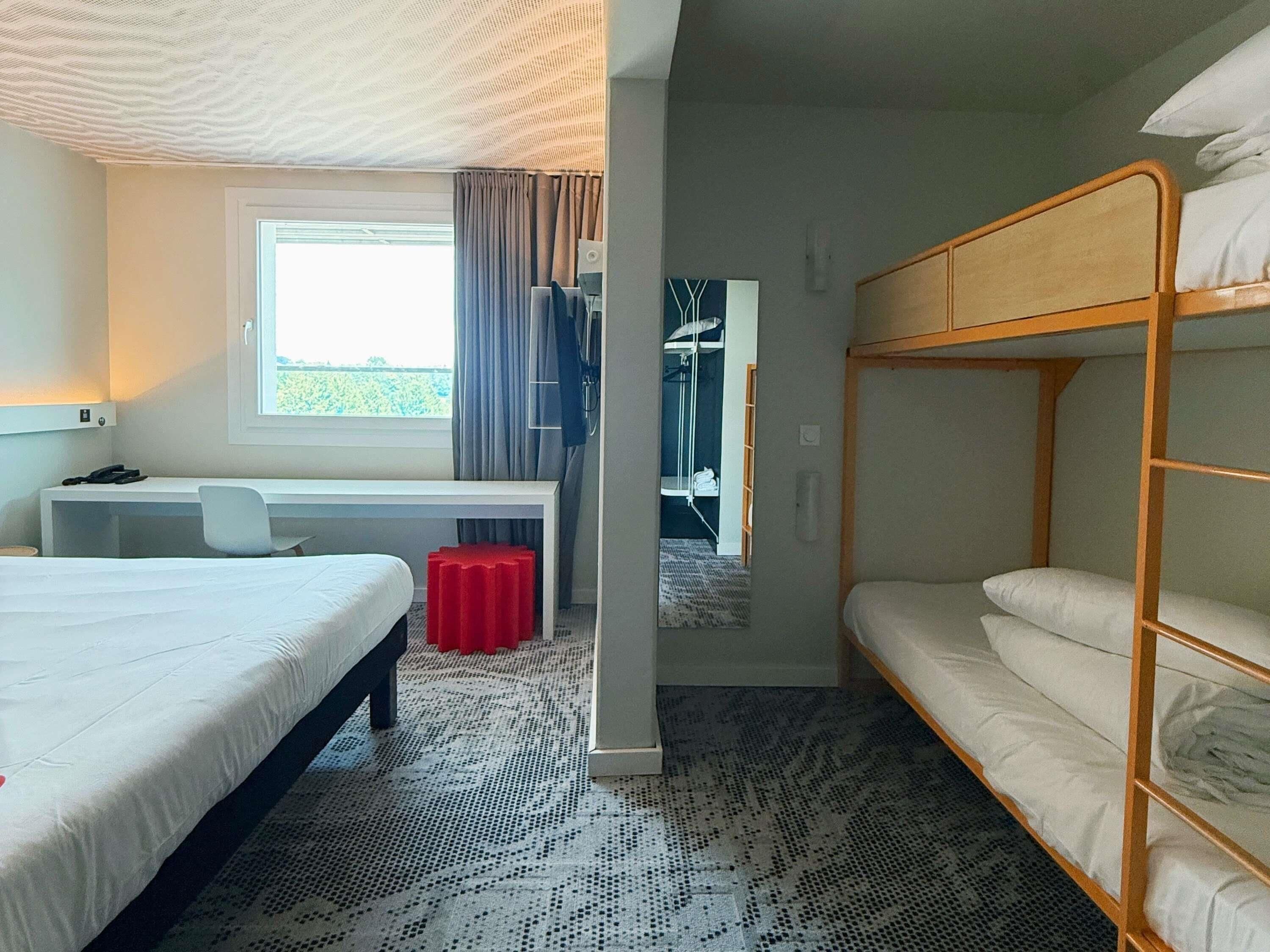 Quarto de hotel com beliche, escrivaninha, cadeira, um banquinho pequeno vermelho e um tapete estampado.