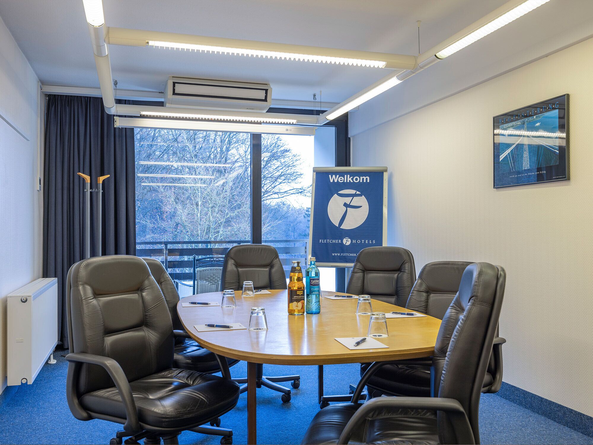 Uma sala de conferências com uma mesa redonda, quatro cadeiras, um letreiro com a palavra 'Welcome' e uma janela com vista para árvores.