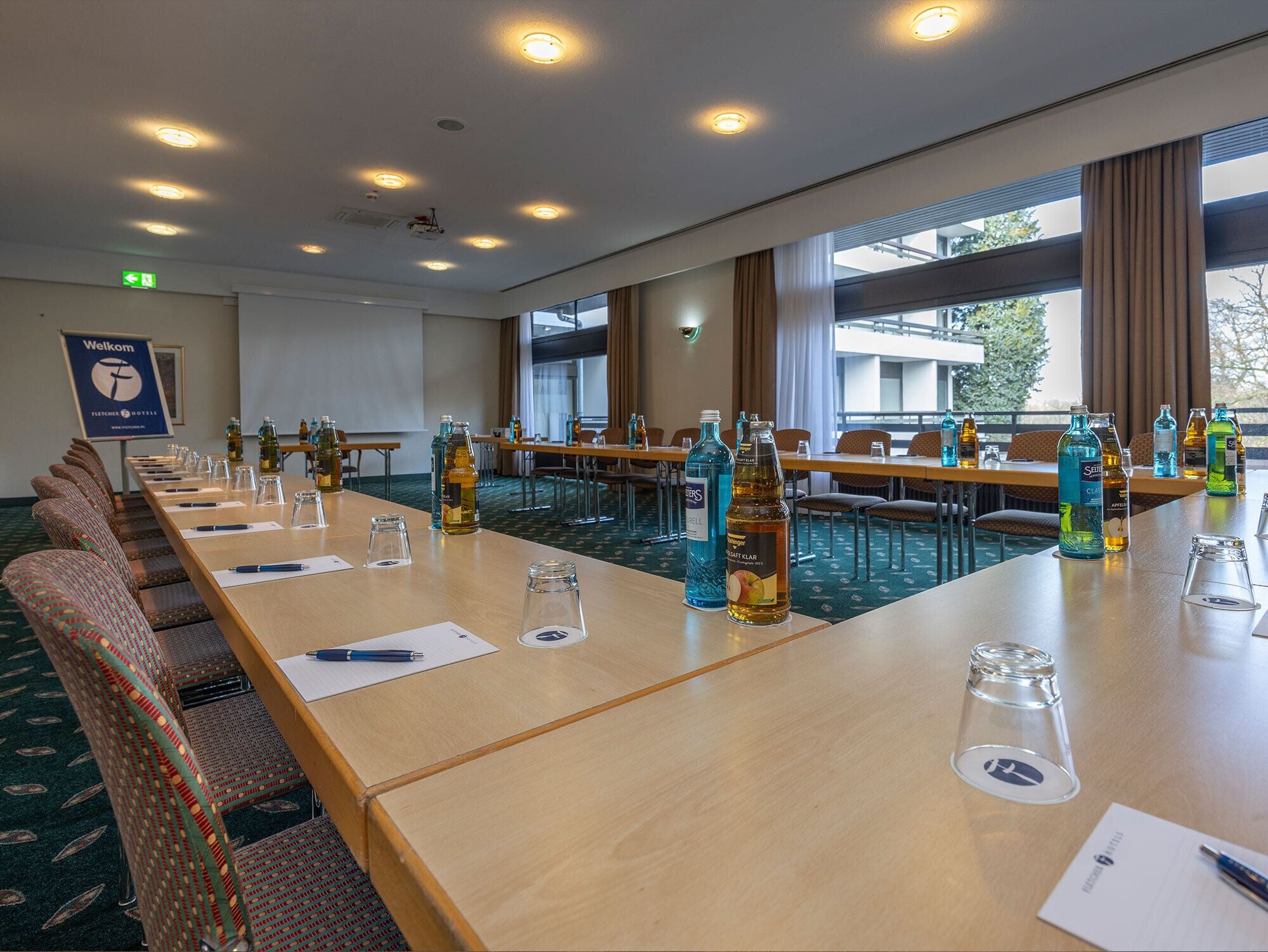 Sala de conferências com mesas compridas, cadeiras e diversas bebidas.