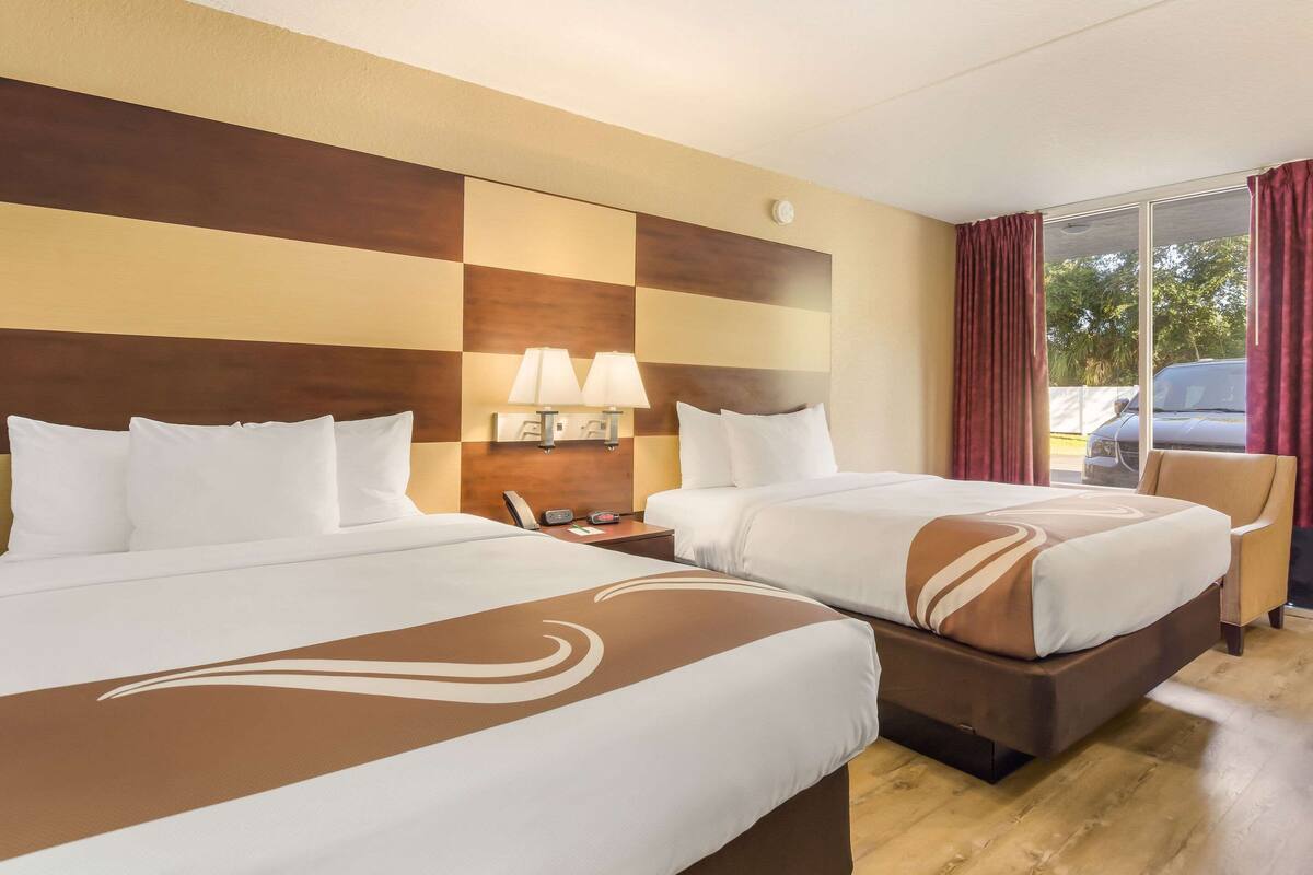 Quarto de hotel com duas camas, uma mesa de cabeceira com abajur, uma cadeira e vista para o exterior.