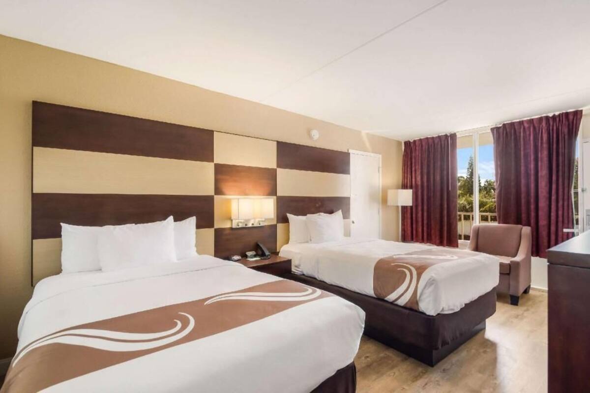Quarto de hotel com duas camas, uma escrivaninha, uma cadeira e vista para o exterior.