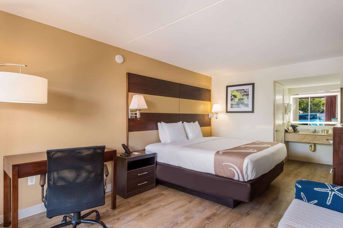 Quarto de hotel com cama grande, escrivaninha com cadeira, mesa de cabeceira, espelho e janela com vista para o exterior.