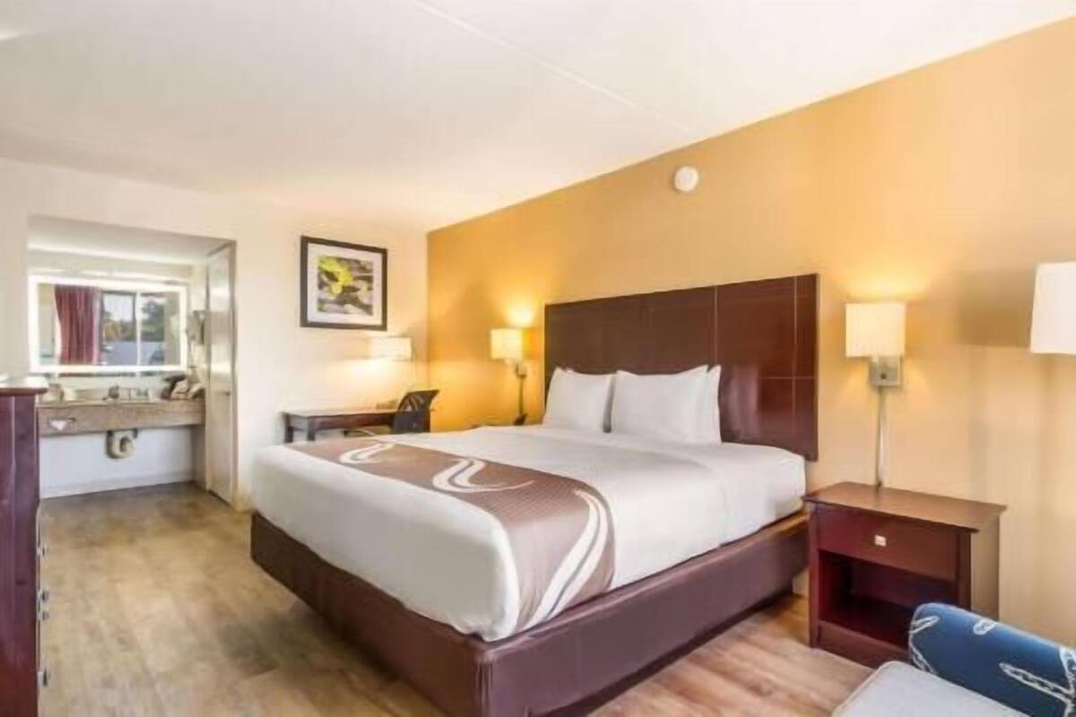 Quarto de hotel com cama, mesa de cabeceira, abajur e espelho.