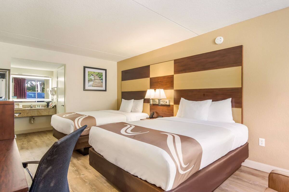 Quarto de hotel com duas camas, uma escrivaninha, uma cadeira, banheiro e janela.