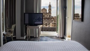 Quarto de hotel com cama, televisão, cadeira e vista para um edifício histórico através de uma porta aberta.