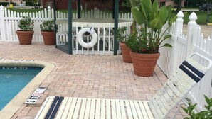 Uma área à beira da piscina com um gazebo de palha, cadeiras de lounge brancas e plantas em vasos.