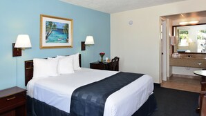 Quarto de hotel com uma cama grande, duas luminárias de cabeceira, um quadro com uma paisagem de praia, uma pequena mesa com um vaso de flores e um banheiro visível através de uma porta aberta.