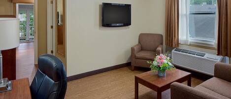 Quarto de hotel com televisão de tela plana, sofá, poltrona, mesinha com um vaso de flores e uma janela grande com cortinas.