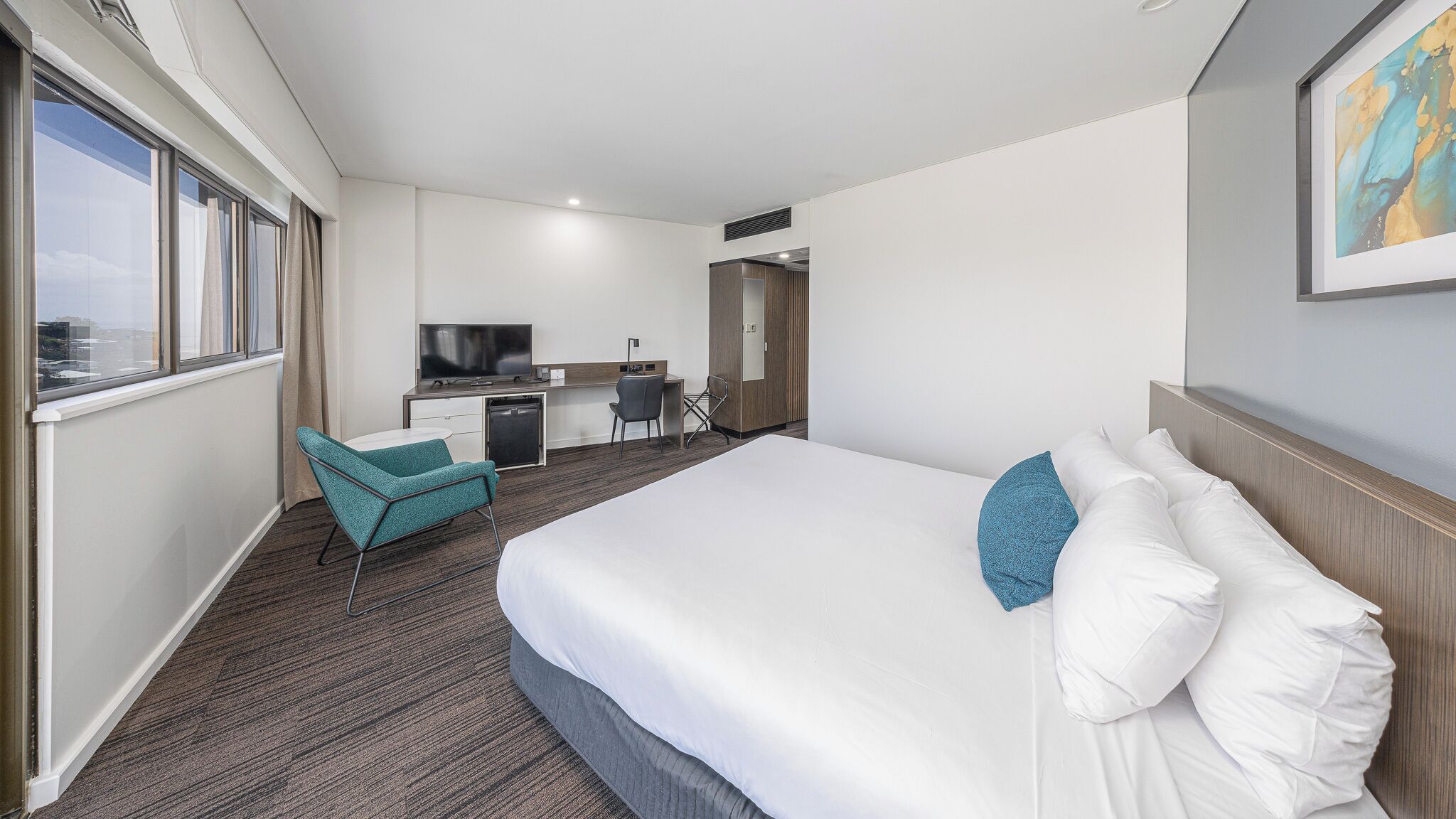 Quarto de hotel com uma cama grande, uma escrivaninha com televisão, uma cadeira e vista pela janela.