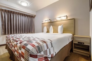 Quarto de hotel com uma cama grande, uma cabeceira de madeira, uma mesa de cabeceira com gaveta e uma janela com cortinas.