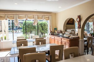 Espaço de refeições de hotel com mesas, cadeiras, um balcão de buffet e grandes janelas com cortinas.