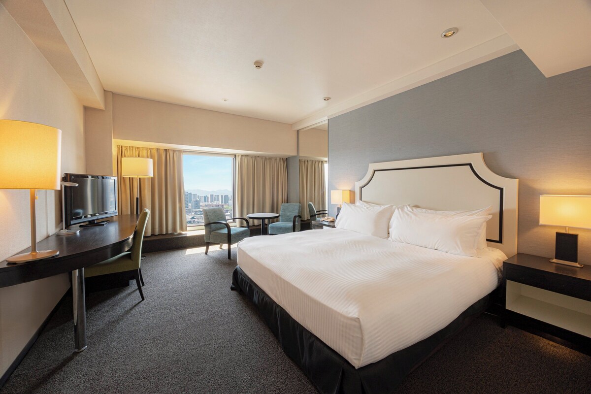 Quarto de hotel com uma cama grande, uma escrivaninha com televisão, uma cadeira, uma mesinha e vista para a cidade pela janela.