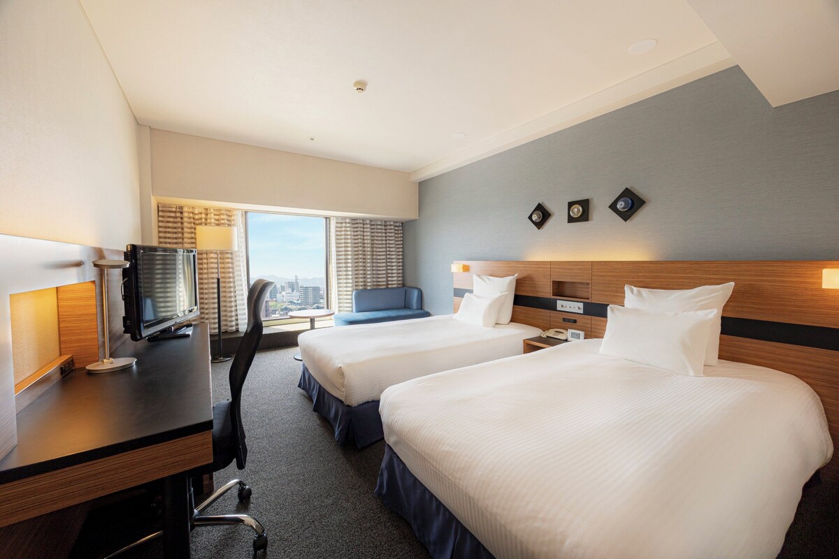Quarto de hotel com duas camas, uma escrivaninha com computador, uma cadeira e vista para a cidade.