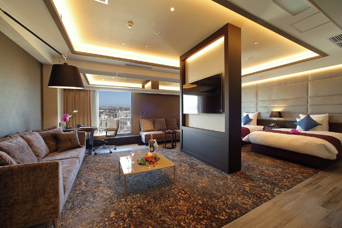 Quarto de hotel moderno com uma cama grande, um sofá, uma mesa de centro com frutas e vista para a cidade.