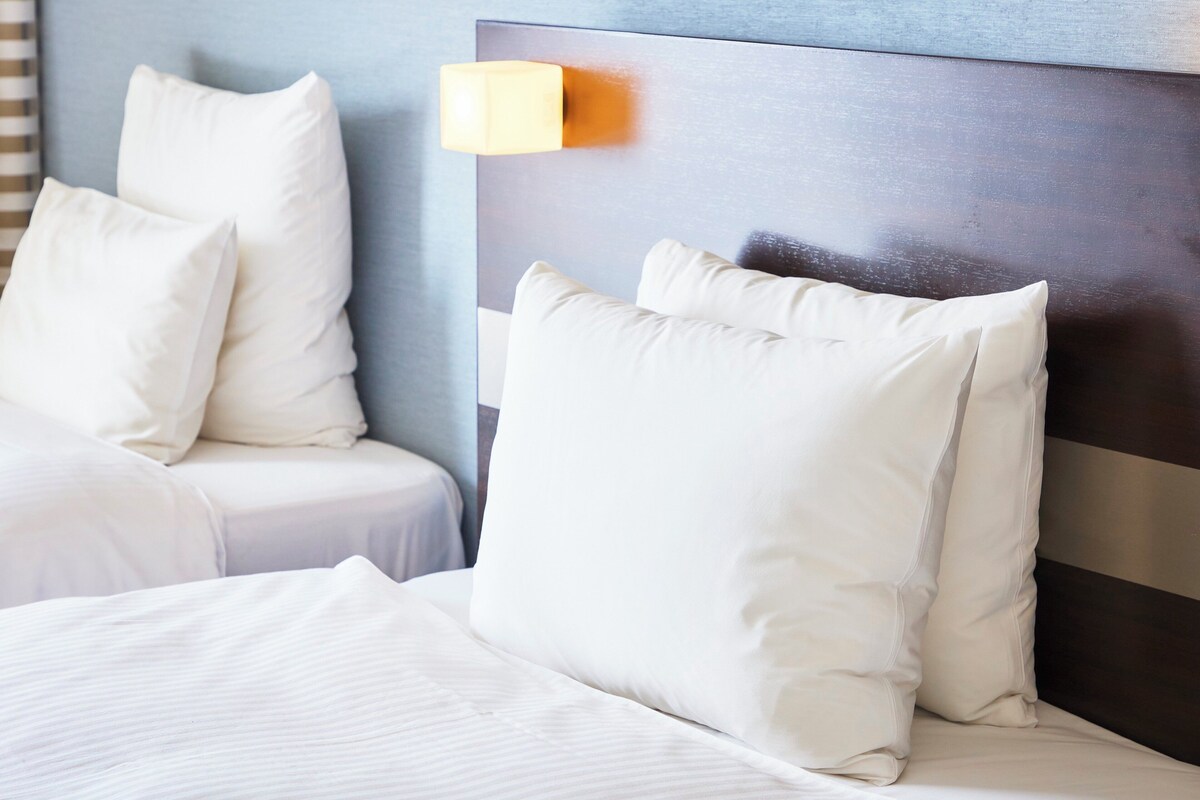 Quarto de hotel com cama, almofadas e luminária fixada na parede.