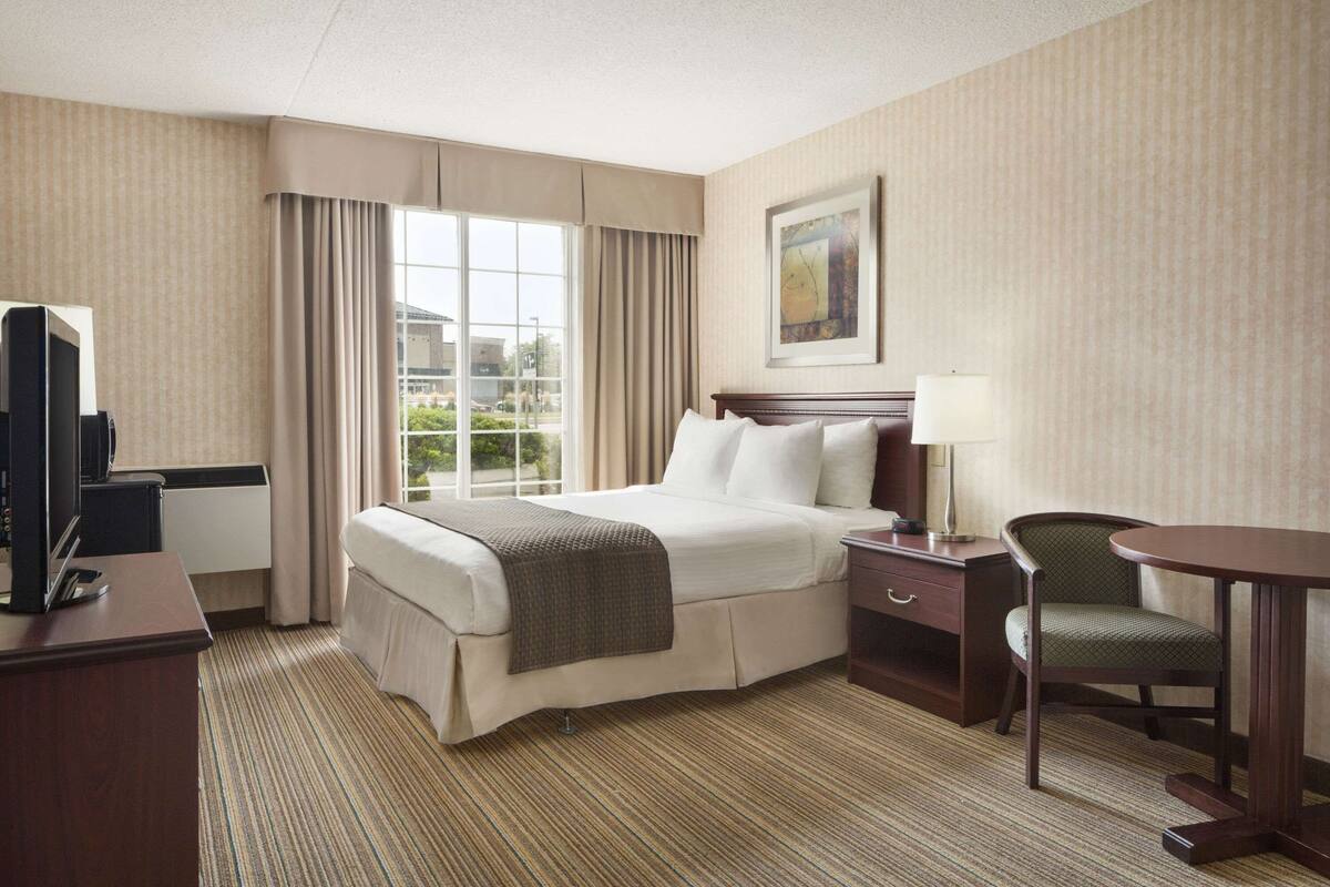 Quarto de hotel com uma cama grande, uma mesa com televisão, uma cadeira, um abajur e uma janela com cortinas.