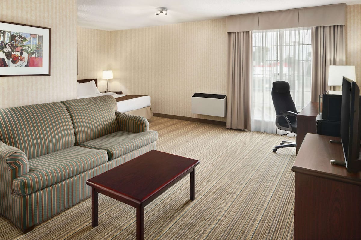 Quarto de hotel com sofá listrado, mesa de centro de madeira, cama com roupa de cama branca, escrivaninha com computador e janela com cortinas.