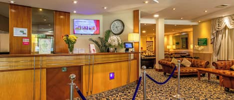 Uma área de recepção de hotel com balcão de madeira, relógio, tela de TV e área de estar com sofás de couro.