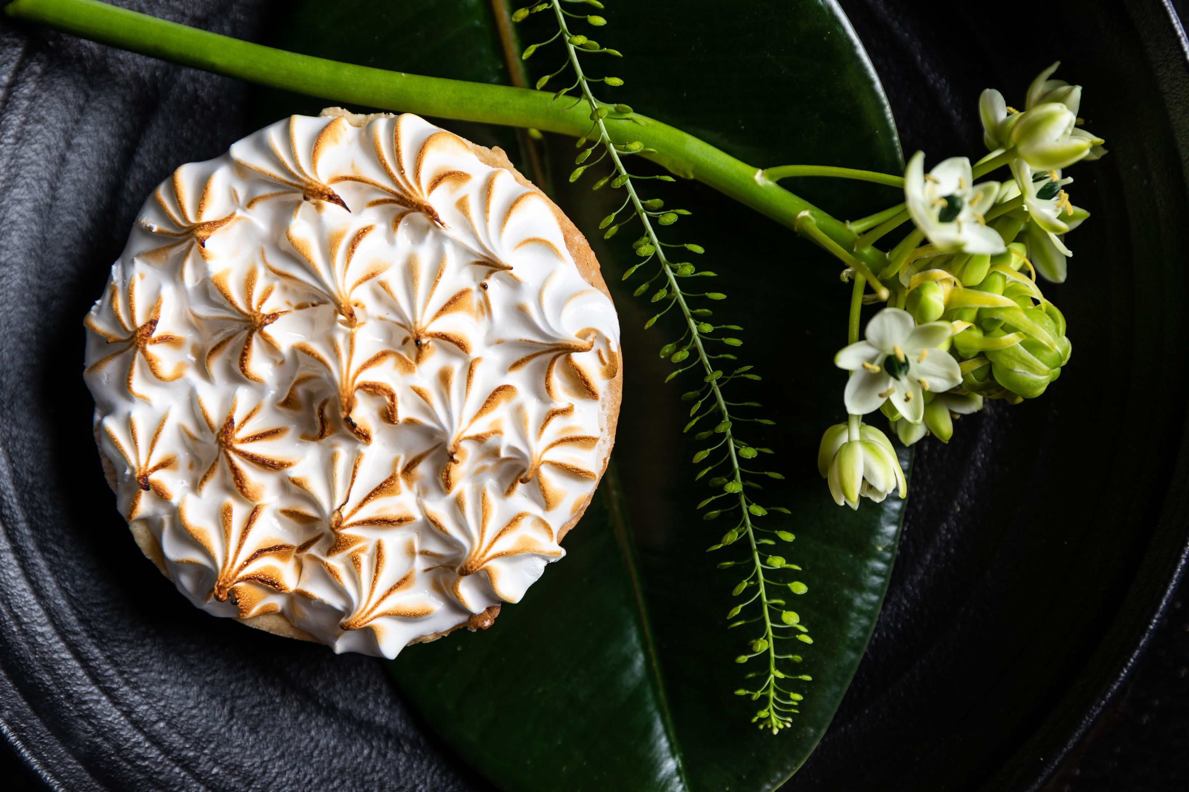 Sobremesa com um padrão decorativo sobre um prato escuro, acompanhada por uma folha verde e flores brancas.