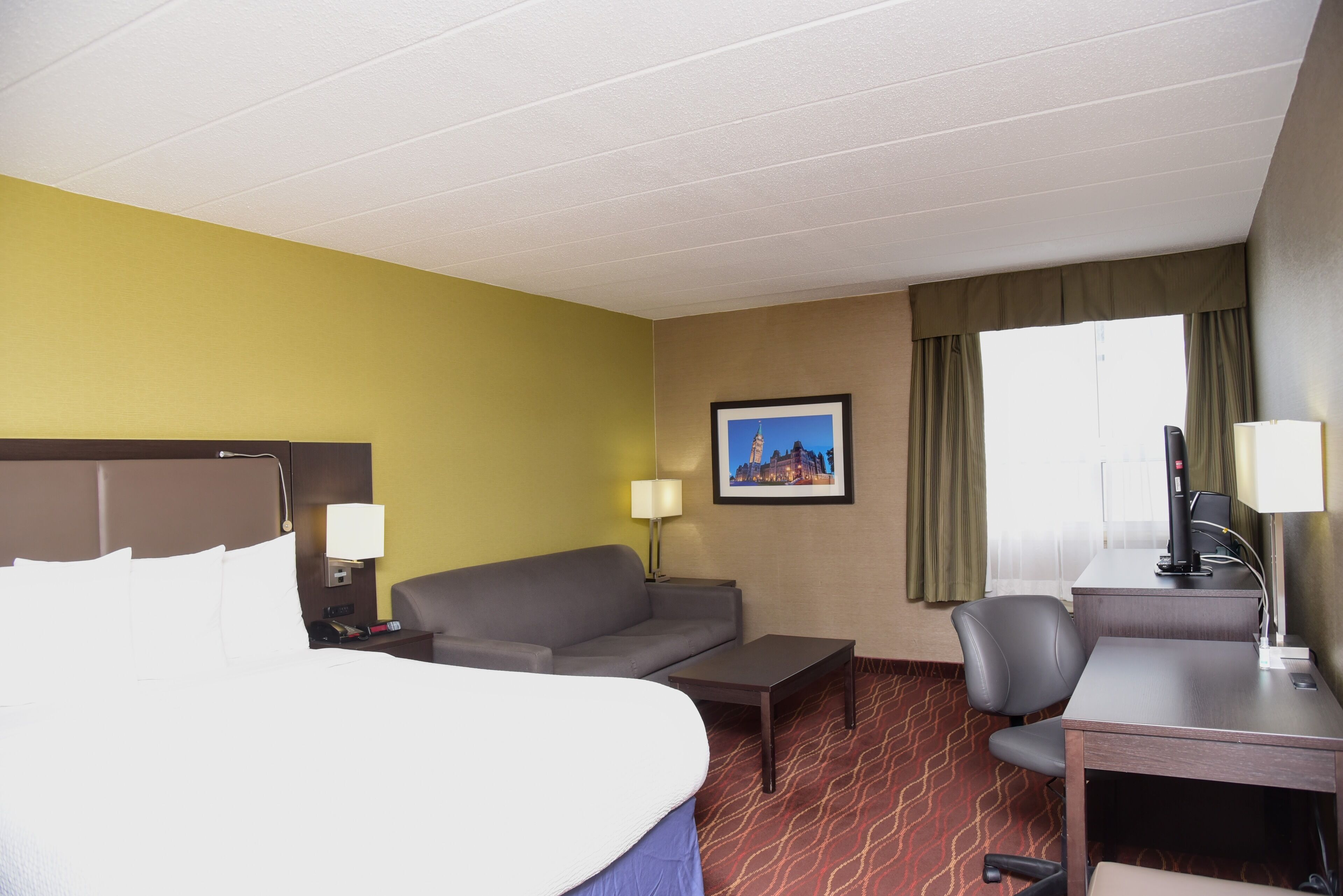 Quarto de hotel com uma cama, um sofá, uma escrivaninha e uma cadeira. Há uma imagem na parede e uma janela com cortinas.