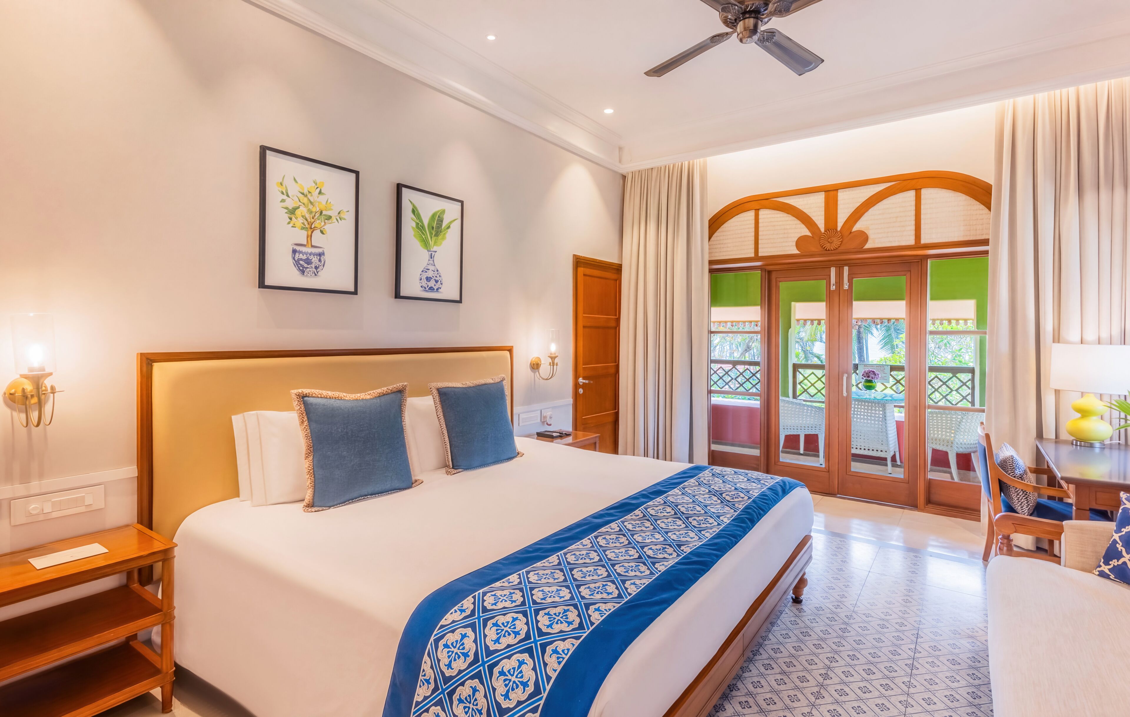 Quarto de hotel com cama grande, cabeceira de madeira, duas luminárias fixadas na parede, ventilador de teto e vista para o exterior através de uma porta de vidro.