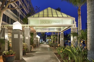 Entrada de hotel com galeria coberta, palmeiras e plantas em vasos.
