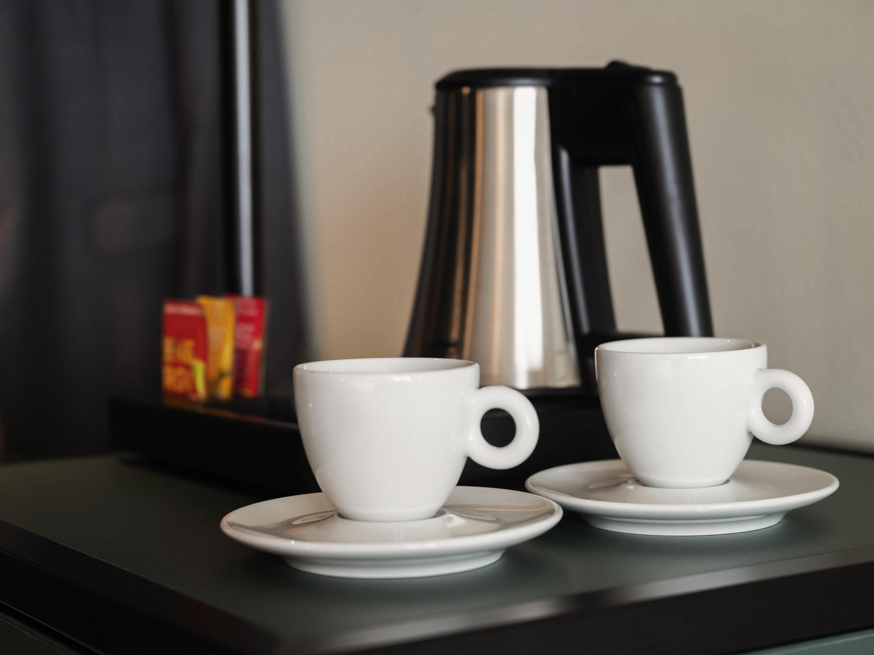 Duas chávenas brancas em pires sobre uma mesa, com uma máquina de café preta ao fundo.