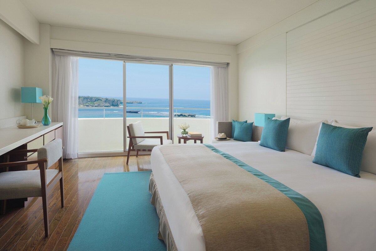 Quarto de hotel com uma cama grande, uma escrivaninha, uma cadeira e vista para o oceano.