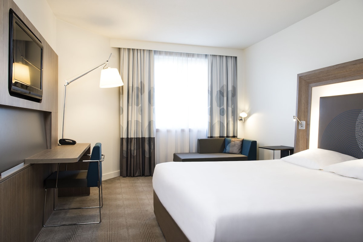 Quarto de hotel com cama, escrivaninha, cadeira, luminária e janela com cortinas.