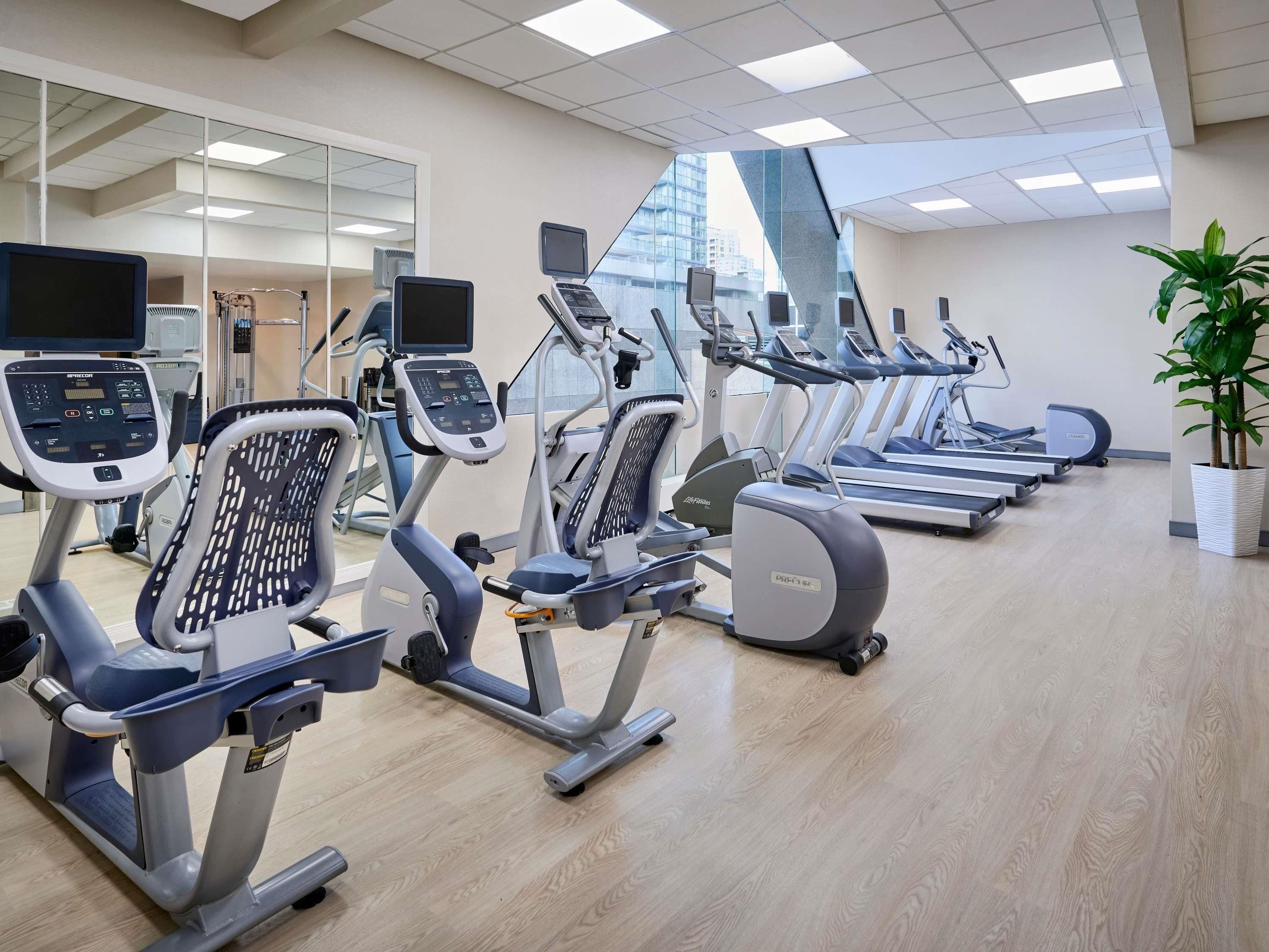 Uma academia com várias máquinas de cardio, um espelho grande e uma planta em vaso.
