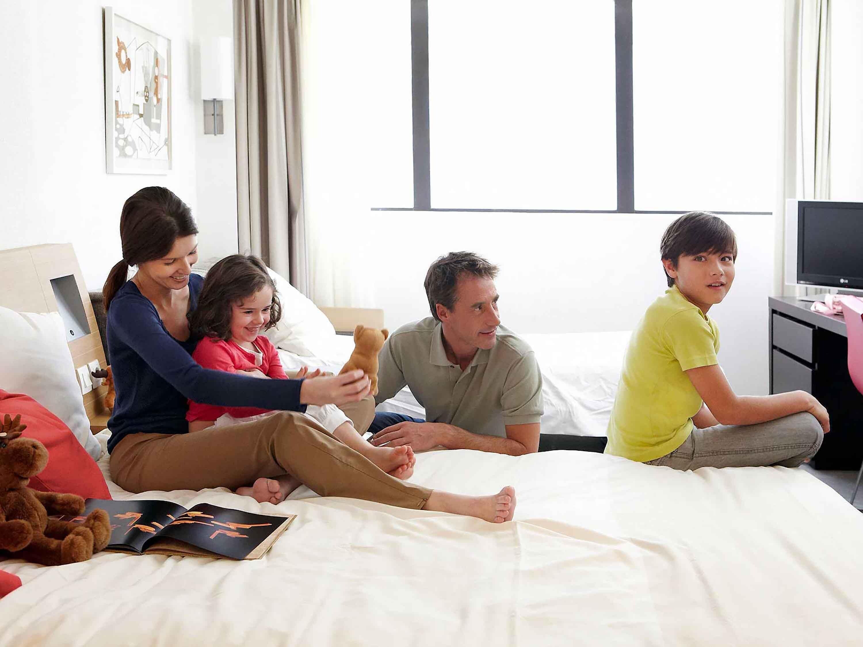 Uma família sentada na cama de um quarto de hotel, com televisão e escrivaninha.