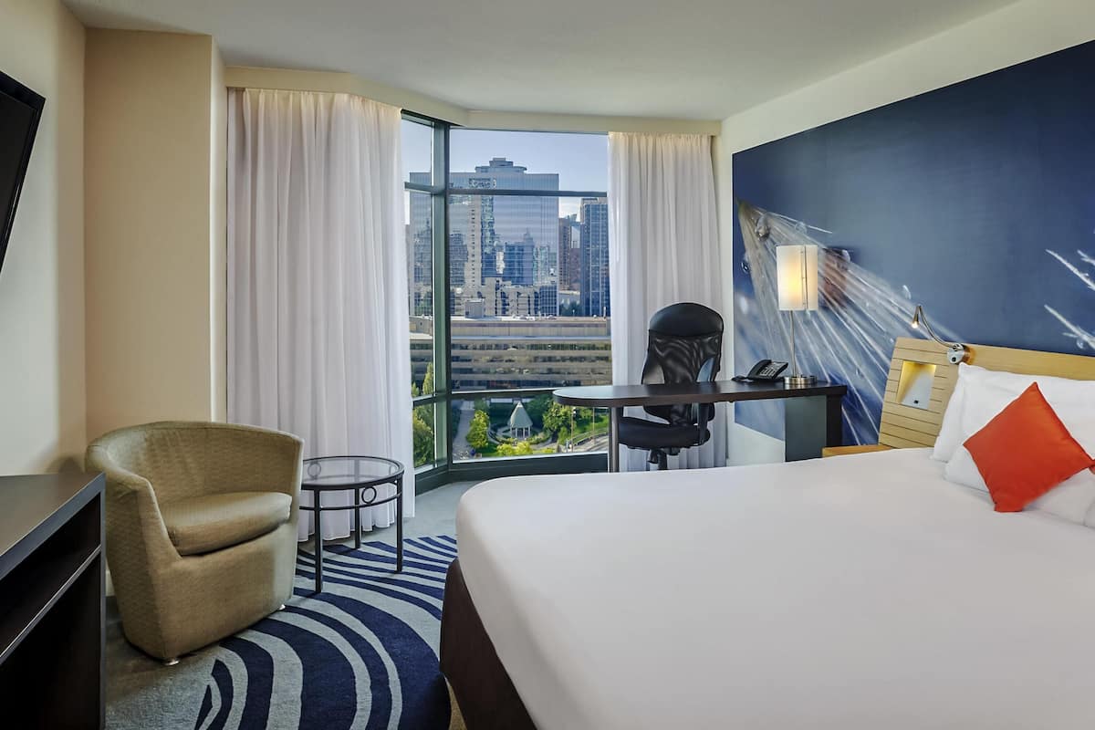 Quarto de hotel com uma cama, escrivaninha, poltrona, televisão e vista para a cidade.