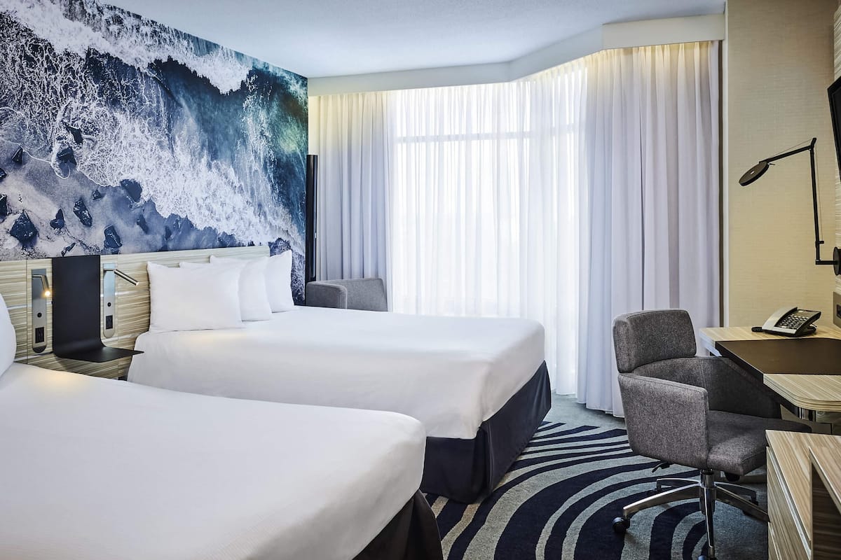 Quarto de hotel com duas camas, uma escrivaninha, uma cadeira, uma janela grande com cortinas e um mural na parede.