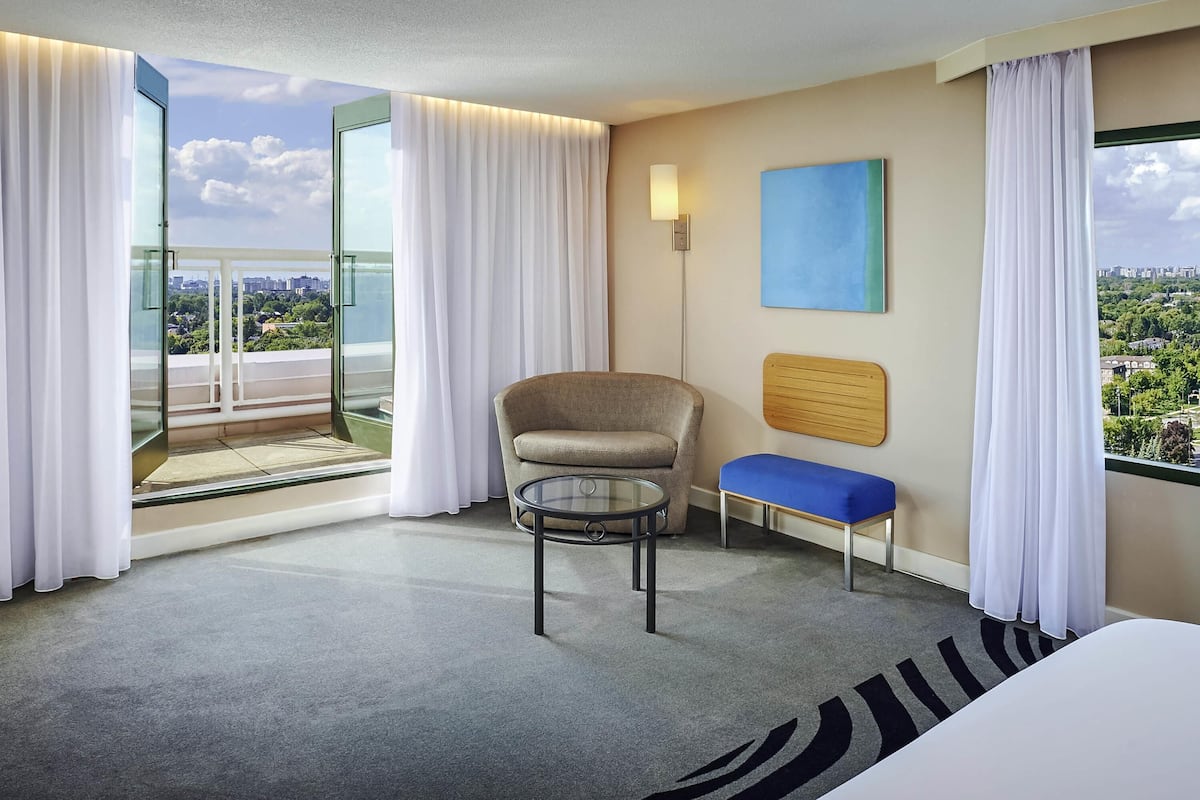 Quarto de hotel com varanda, uma cadeira, uma mesa, um quadro e vista para a cidade.