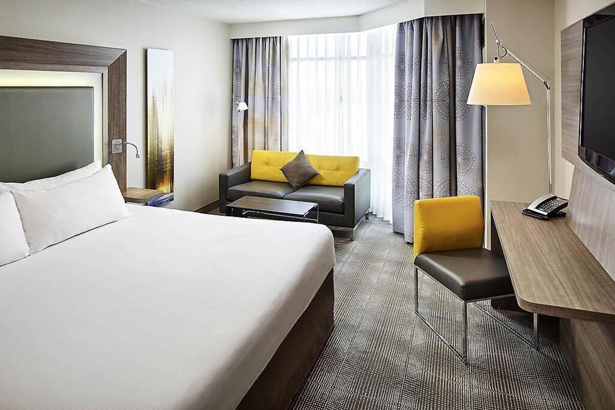 Quarto de hotel com uma cama grande, sofá, escrivaninha, cadeira, televisão e janela com cortinas.