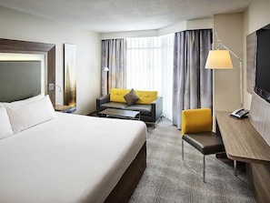 Quarto de hotel com cama, sofá, escrivaninha, cadeira, televisão e janela com cortinas.