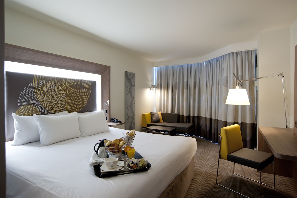 Quarto de hotel com uma cama grande, uma escrivaninha, uma cadeira e uma mesa de jantar posta para o café da manhã.