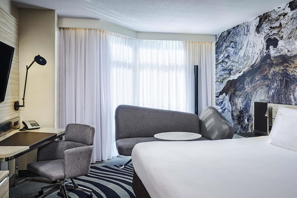 Quarto de hotel com cama, duas poltronas, escrivaninha com telefone e uma janela grande com cortinas.