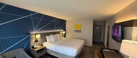 Quarto de hotel com cama, escrivaninha, cadeira, televisão e parede com padrão geométrico.