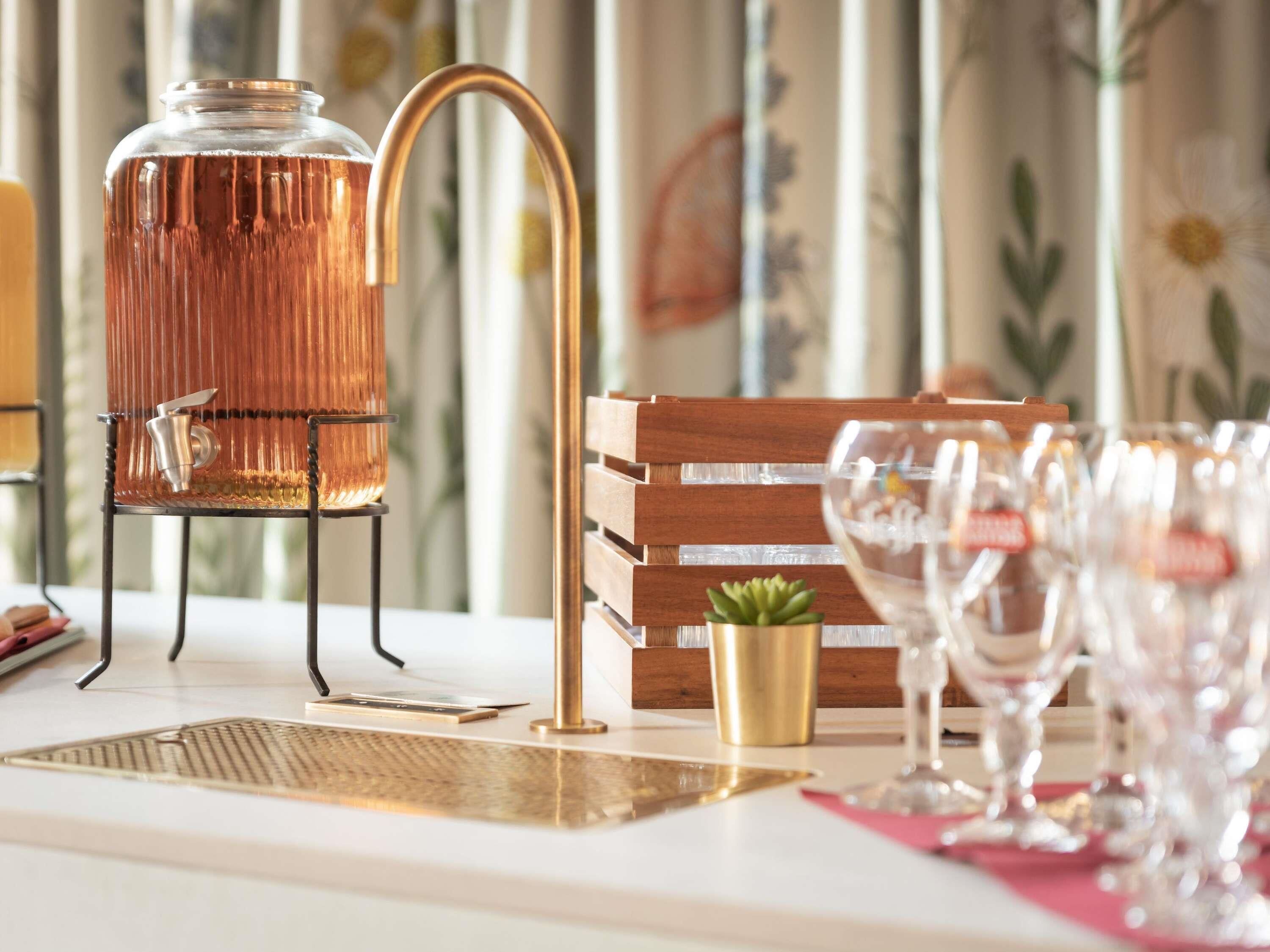 Uma área de bar com um recipiente de vidro cor de cobre, uma torneira dourada e um bloco de madeira com copos.