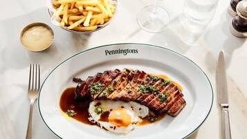 Prato de bife com ovo frito e molho, acompanhado de batatas fritas em uma tigela e um pequeno recipiente com molho, sobre uma mesa branca com um copo e saleiro.