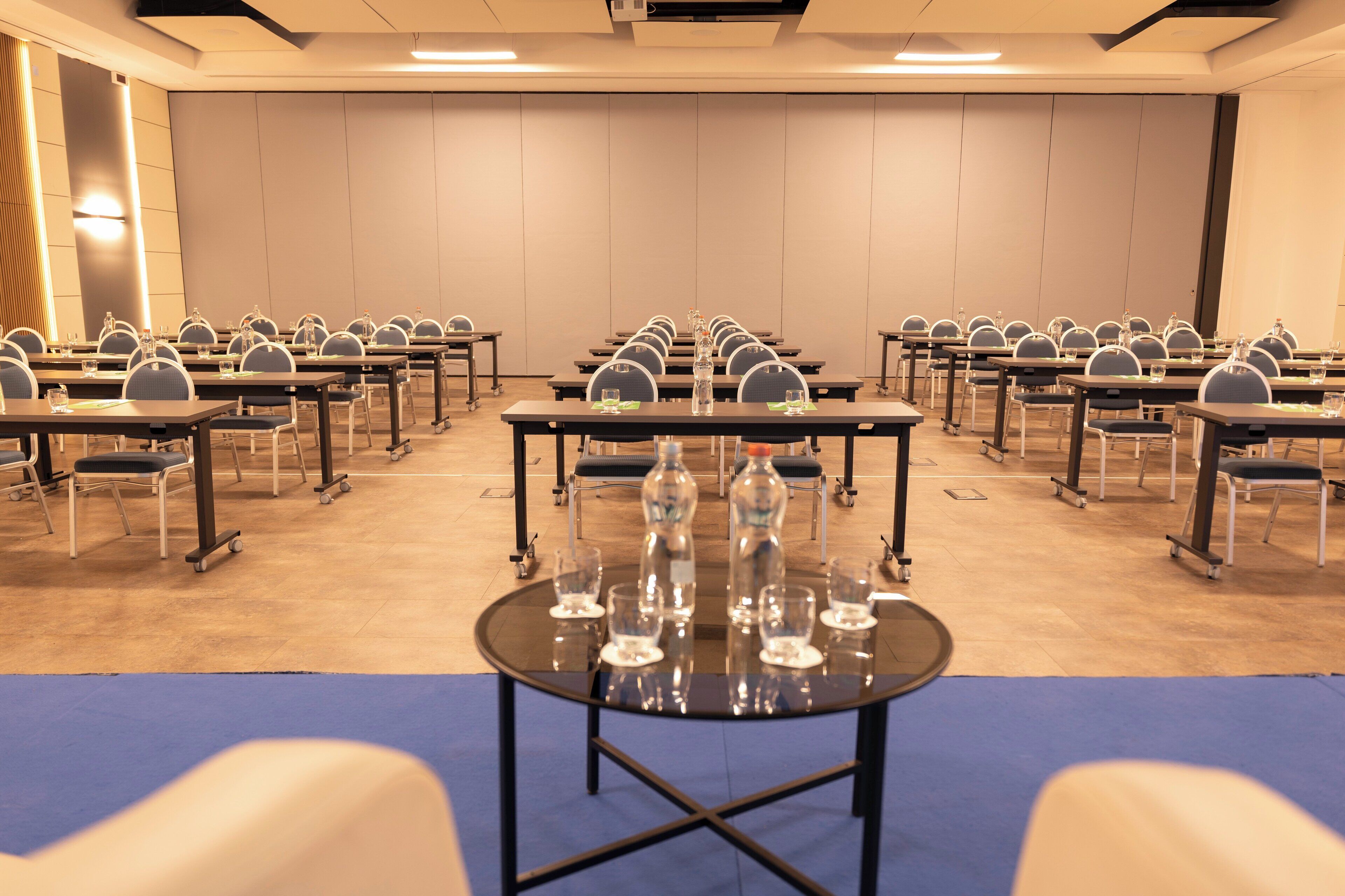 Uma sala de conferências com mesas e cadeiras dispostas em fileiras, equipada com garrafas de água e copos.