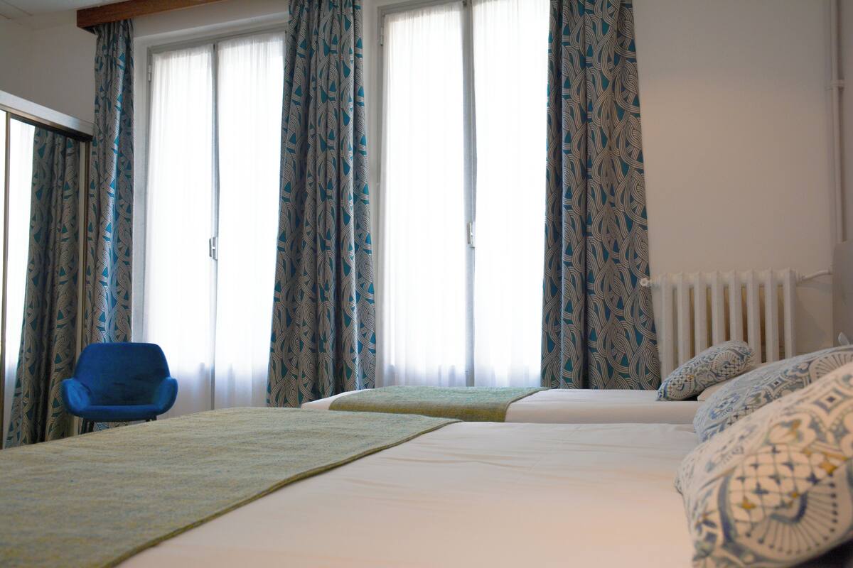 Quarto de hotel com cama, uma cadeira azul, cortinas estampadas e um radiador.