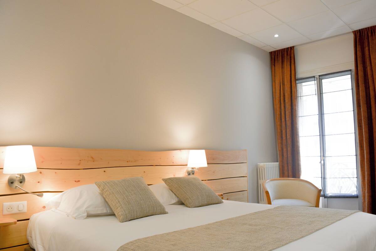 Quarto de hotel com uma cama grande, duas luminárias de cabeceira, um painel de cabeceira de madeira, uma janela com cortinas e uma cadeira.