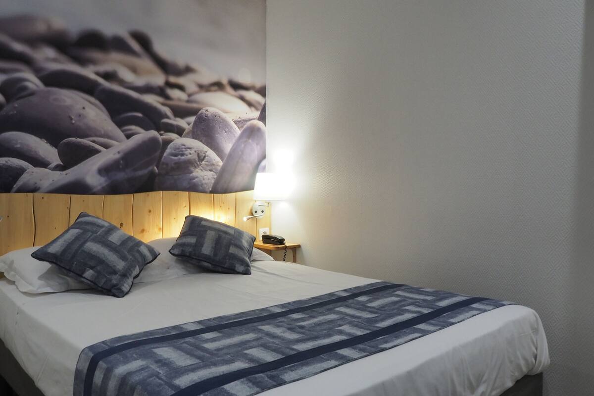 Uma cama com um cobertor estampado, duas almofadas, uma mesa de cabeceira com abajur e um telefone fixado na parede.
