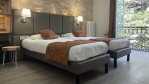 Quarto com parede de pedra, duas camas, uma mesa de cabeceira, um abajur e vista para o verde através de uma varanda.