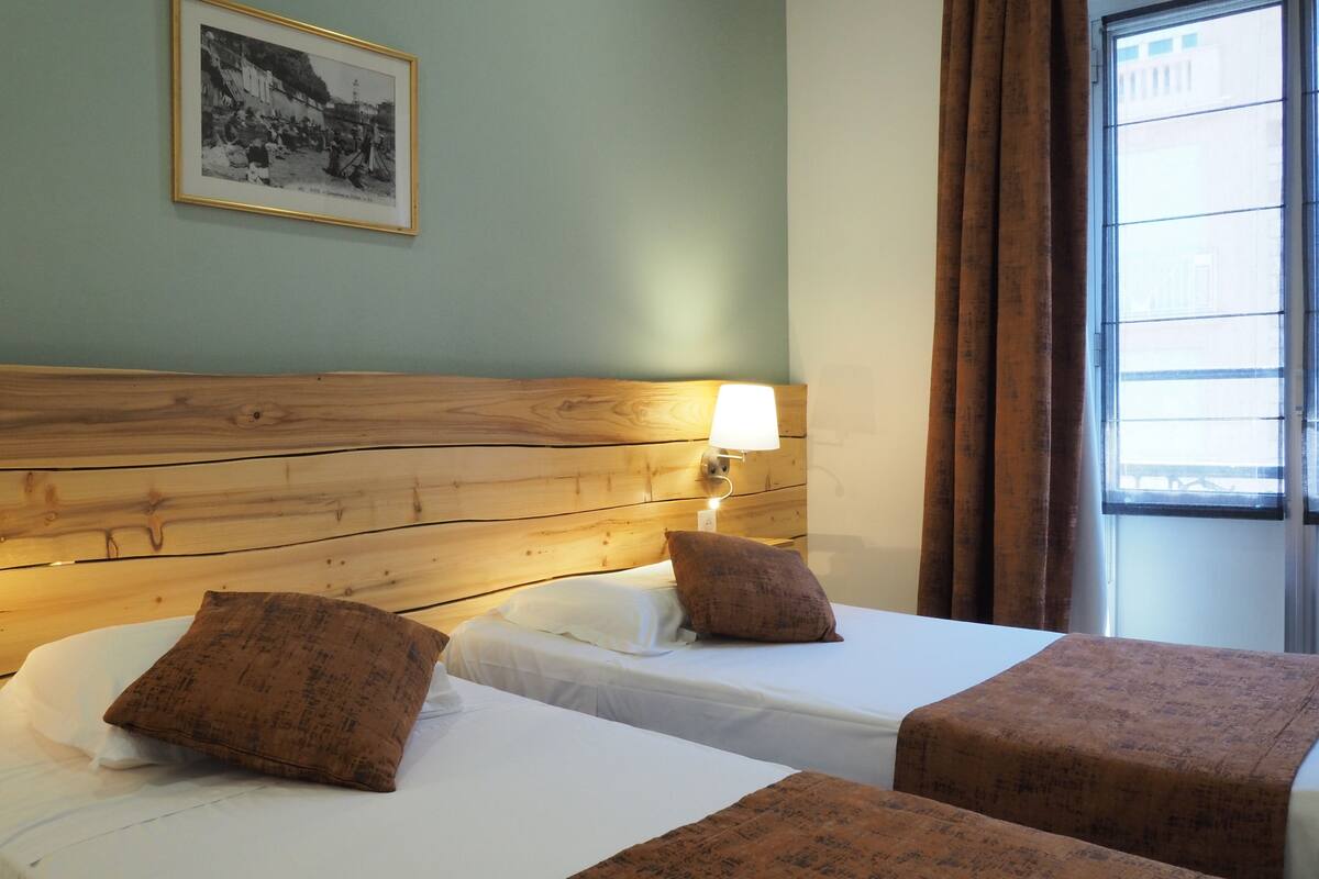 Quarto com duas camas, cabeceira de madeira, quadro pendurado na parede e janela com cortinas.