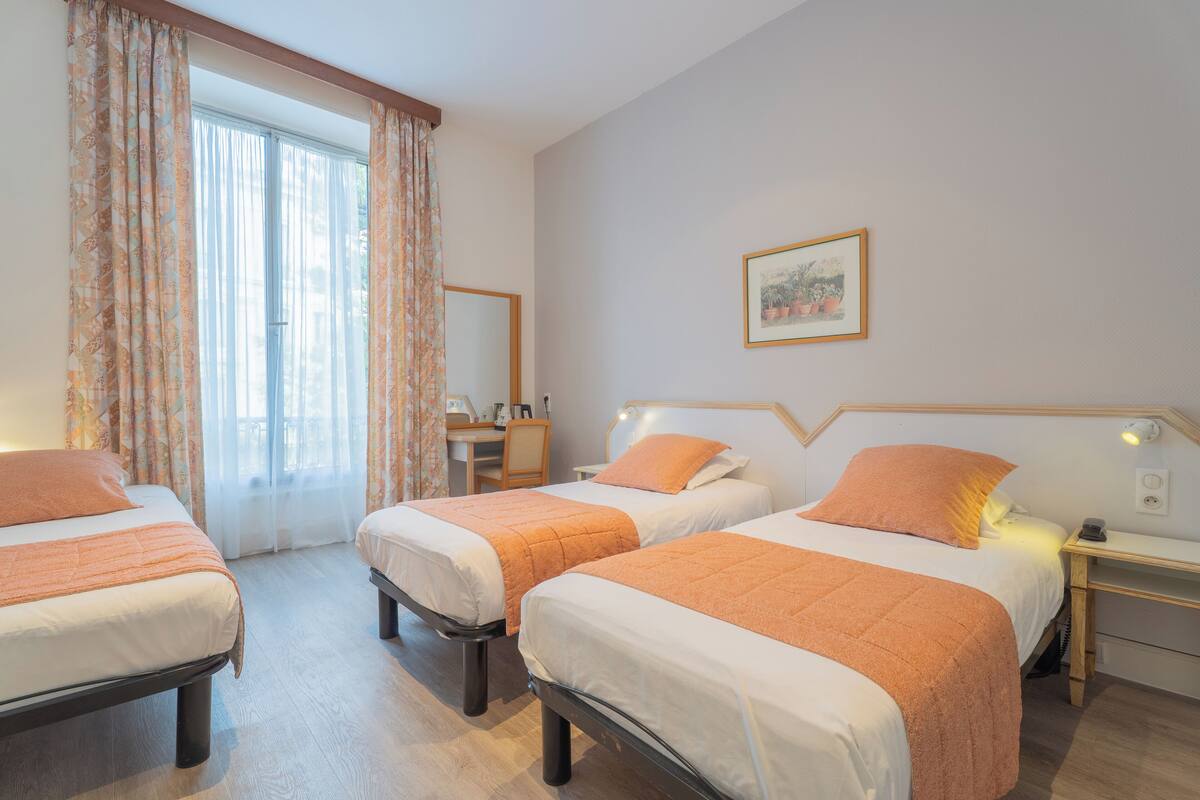 Quarto de hotel com duas camas, uma escrivaninha, uma cadeira, uma janela com cortinas e um quadro na parede.