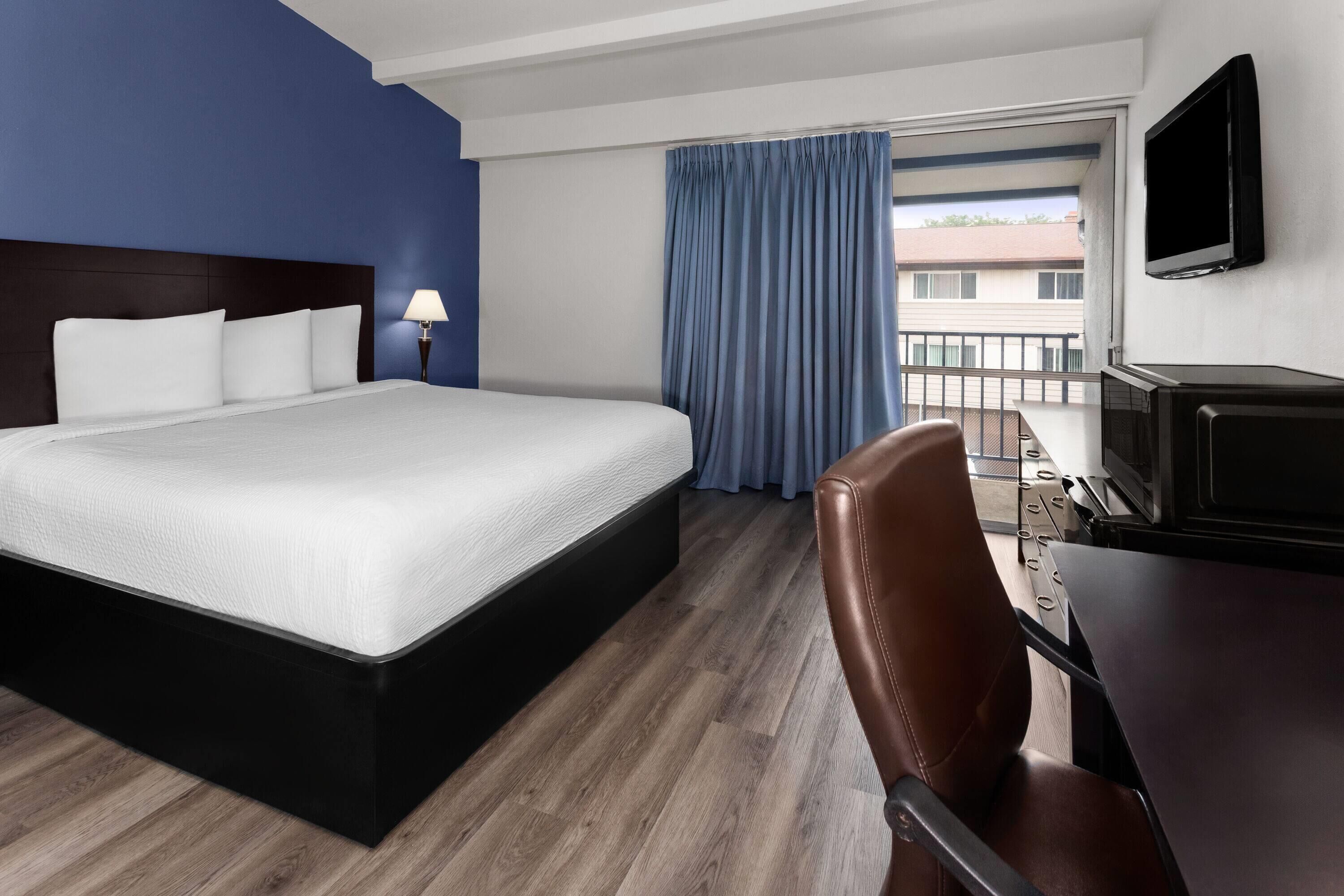 Quarto de hotel com uma cama grande, uma escrivaninha com cadeira, televisão e varanda com vista para edifícios.
