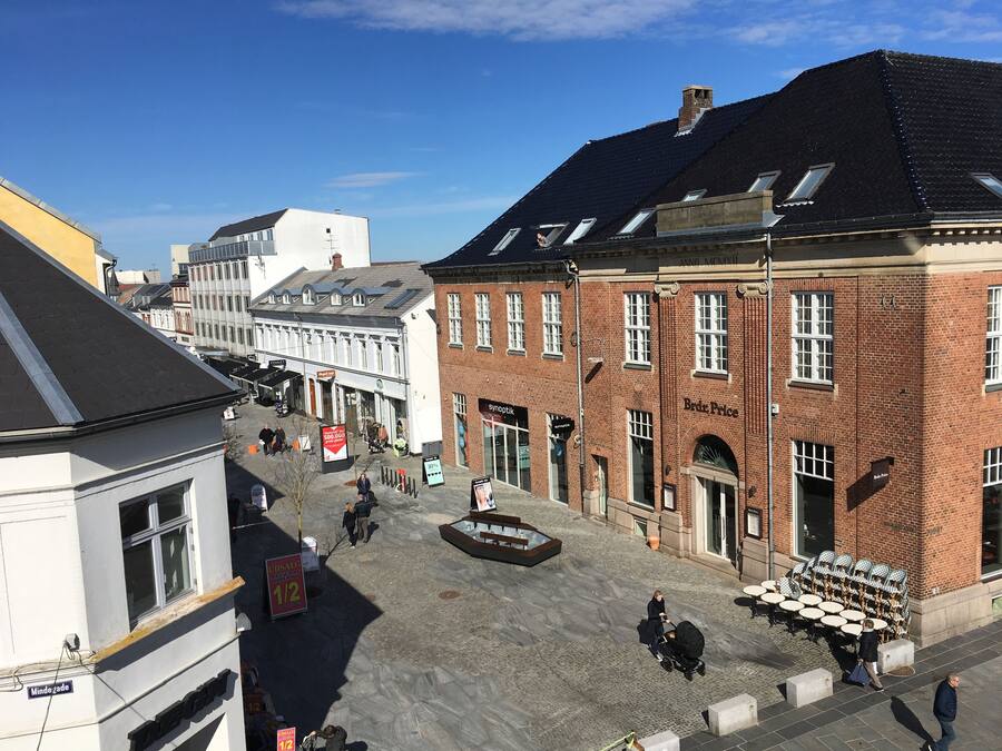 Uma praça de paralelepípedos com edifícios históricos, mesas ao ar livre e uma fonte.